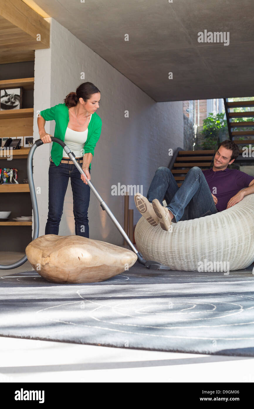 Woman cleaning house with a vacuum cleaner with her husband sitting on ...