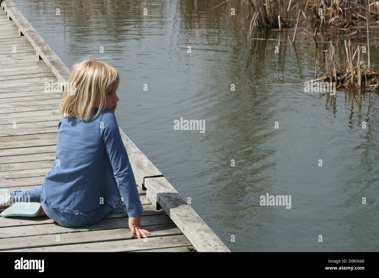 A pretty young blond girl sitting at the edge of a dock on a lake in ...