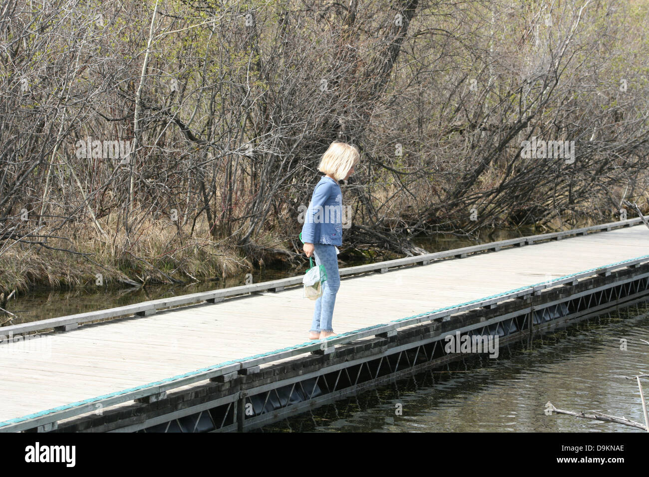 A pretty young blond girl standing on a dock on a lake in spring in ...