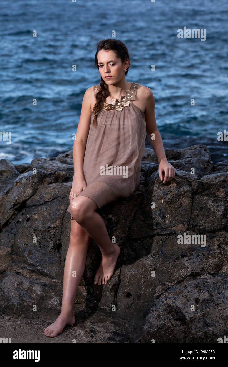 Pretty young woman resting on rocks by the sea looking down pensive ...