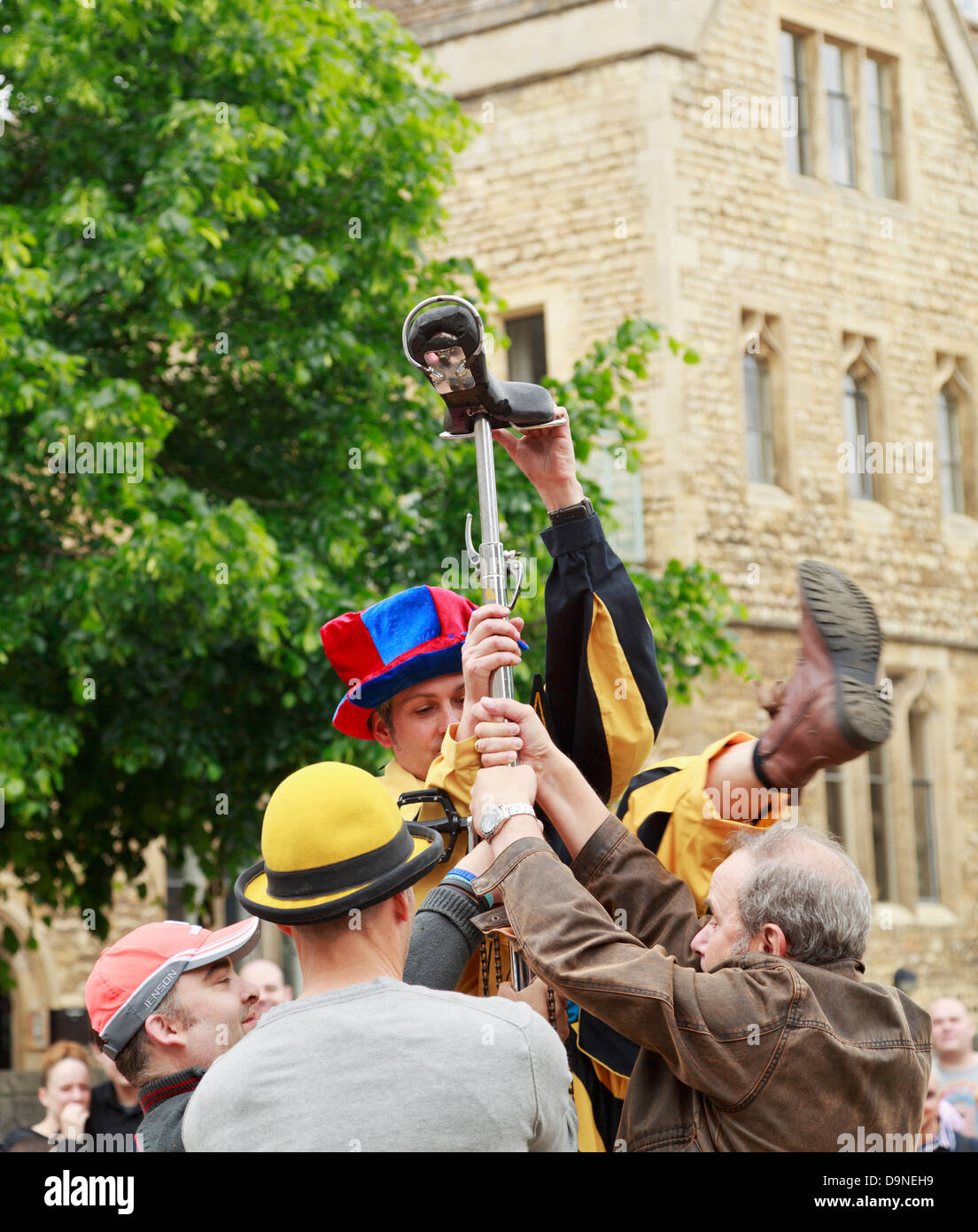 Jester riding a tall unicycle with 3 volunteers helping to balance him ...