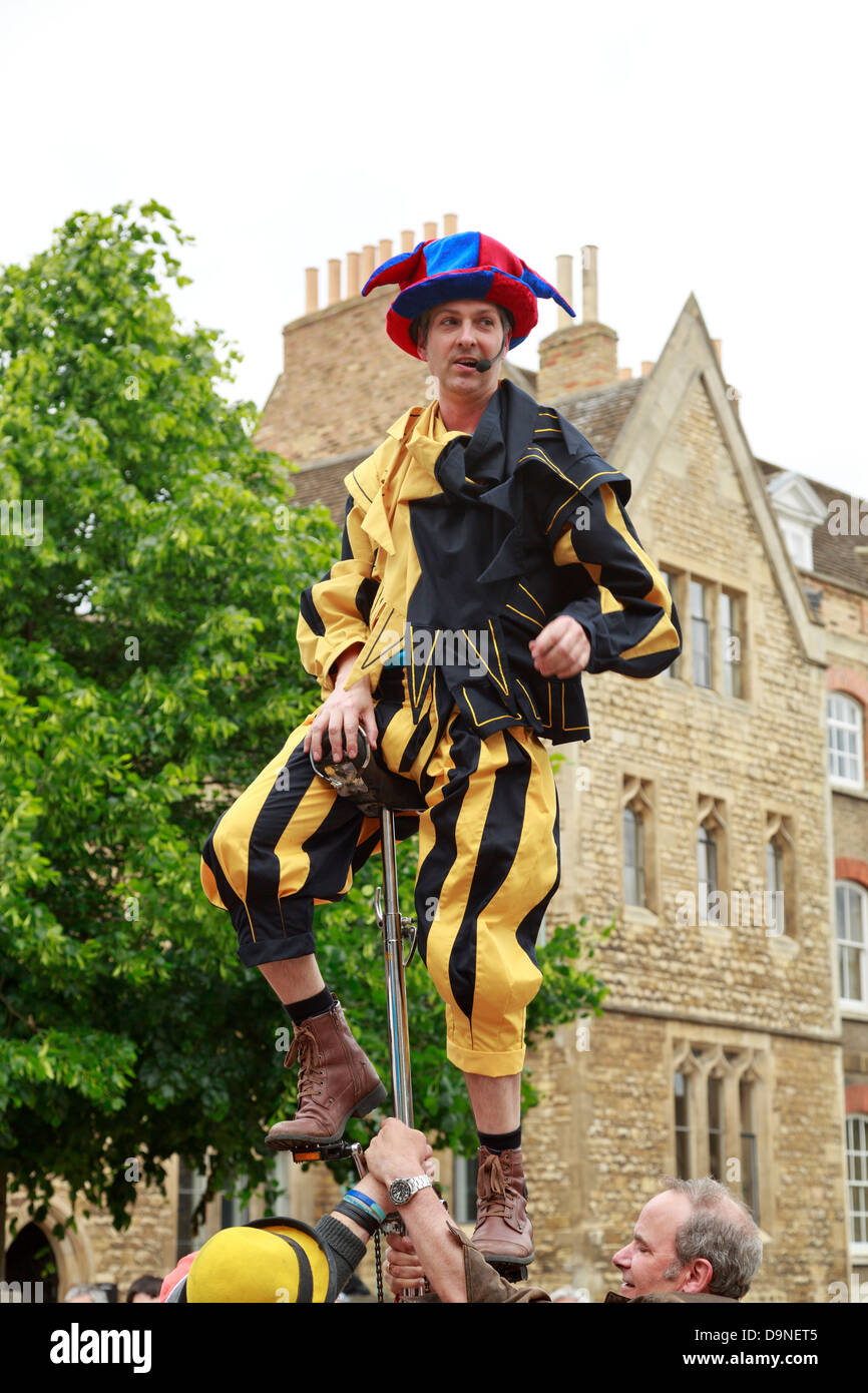 Jester riding a tall unicycle with 3 volunteers helping to balance him ...