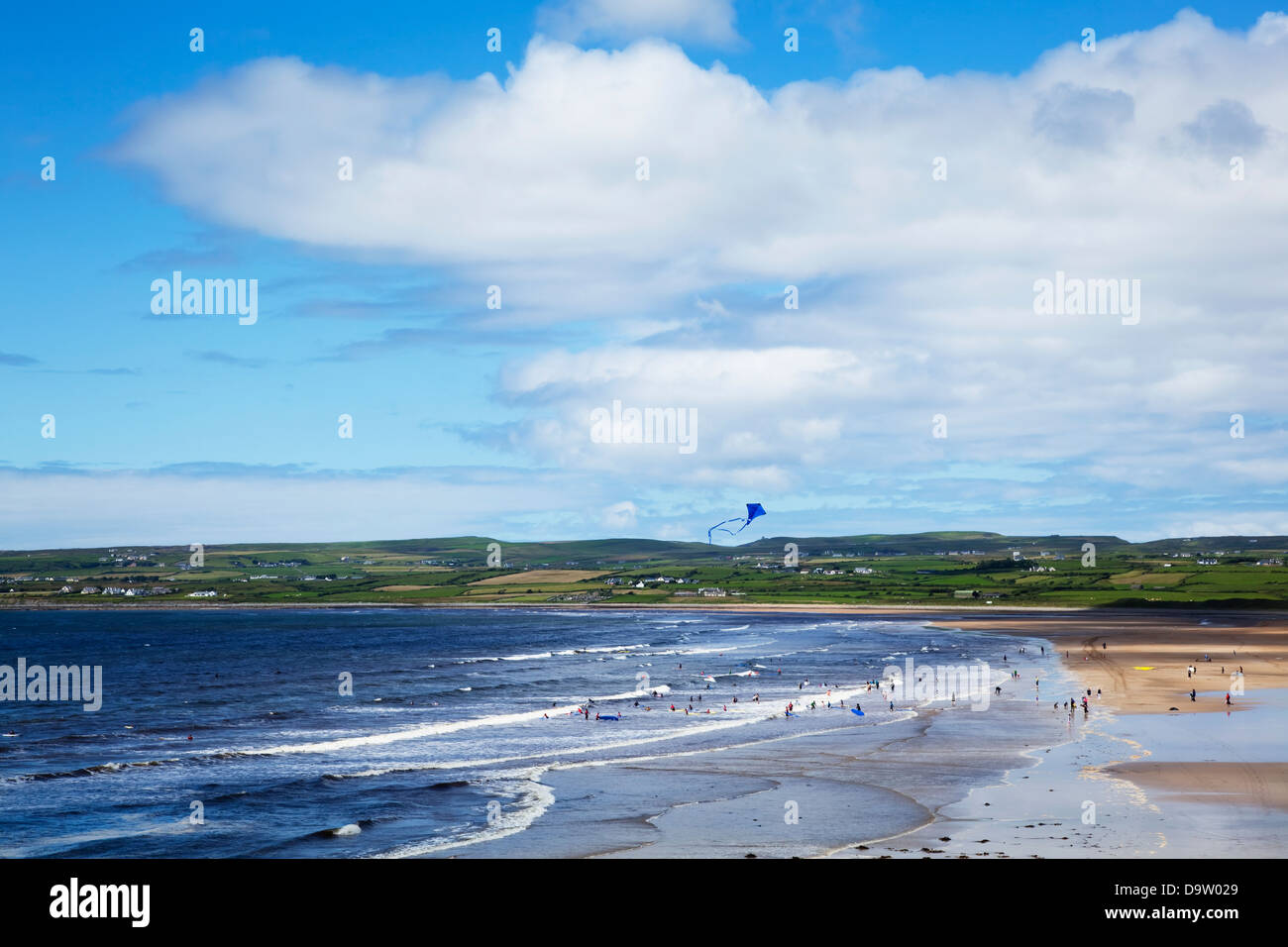 A busy beach with people surfing swimming and flying kites;Lahinch ...