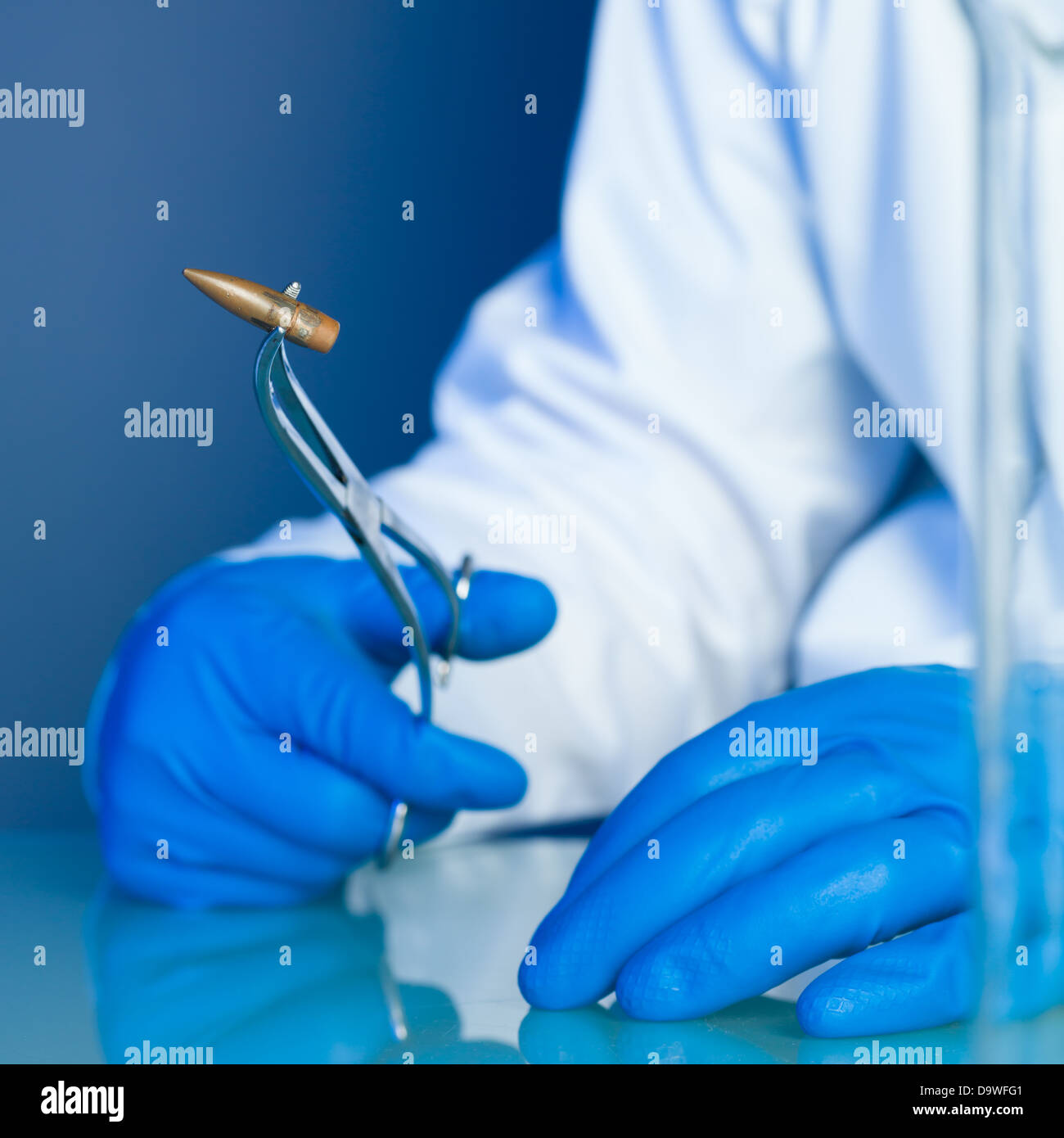 person in a white lab coat and blue rubber gloves, holding a bullet on ...