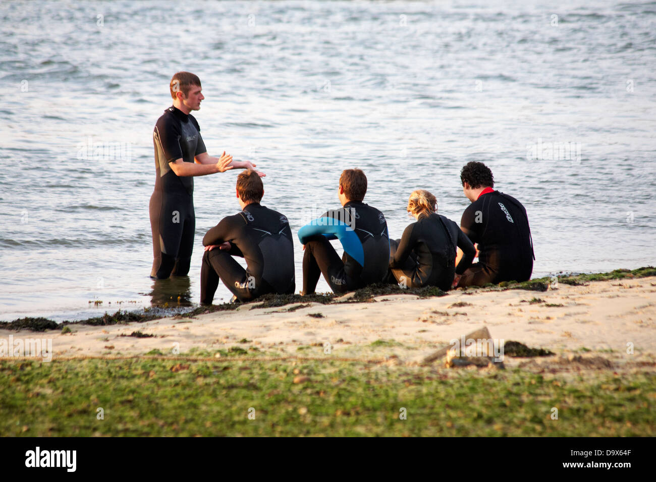 group in wetsuits sat on beach with one standing in water at ...