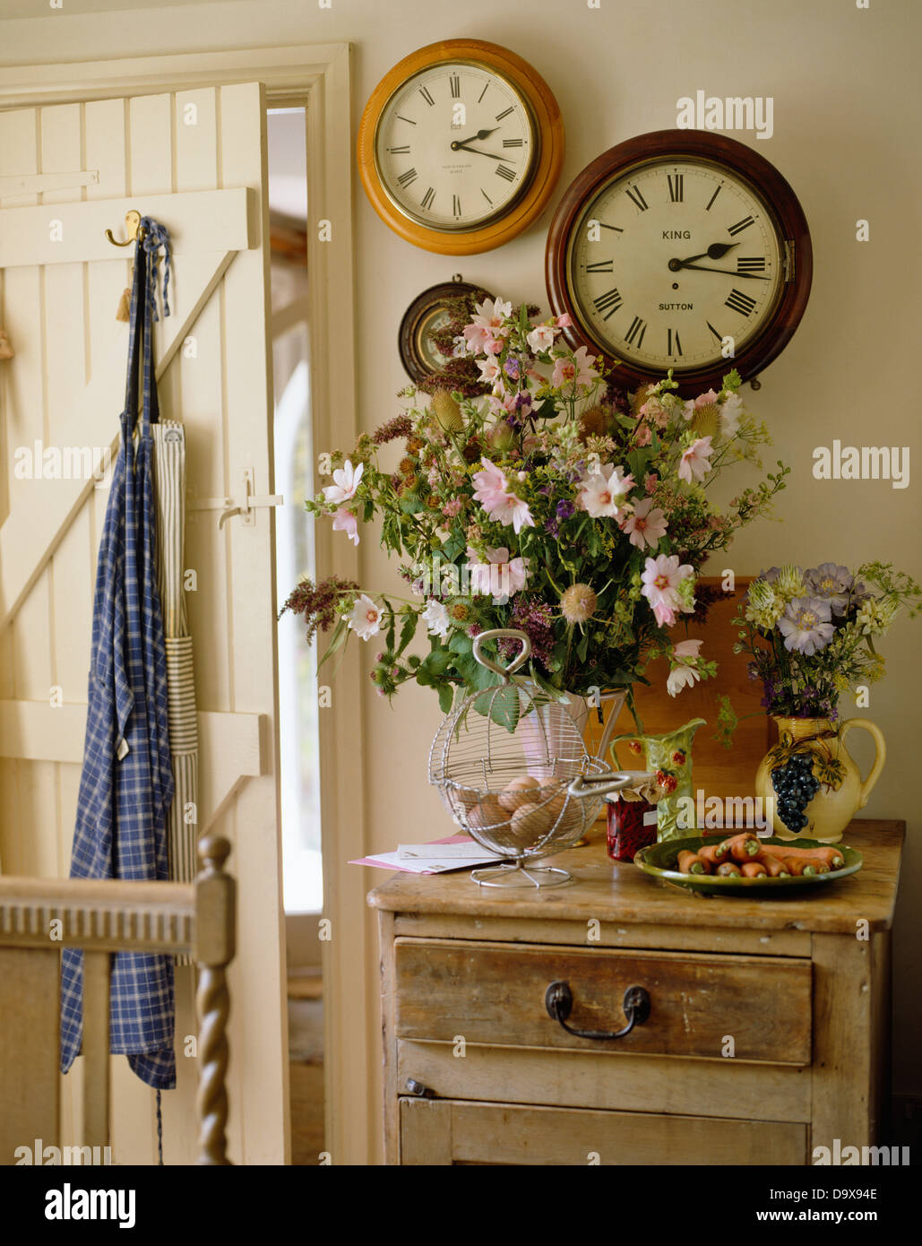 Three circular clocks on wall above rustic cupboard with vase of summer ...