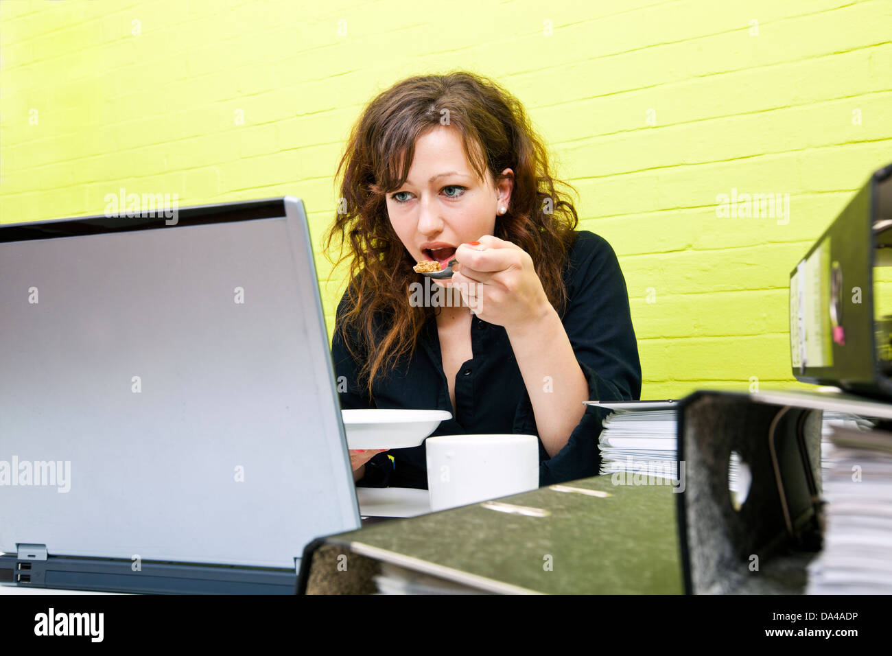 Caucasian young woman eating and working on her laptop computer at her ...
