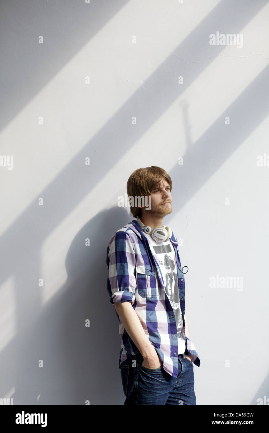 young man leaning against white wall listening to music on headphones ...