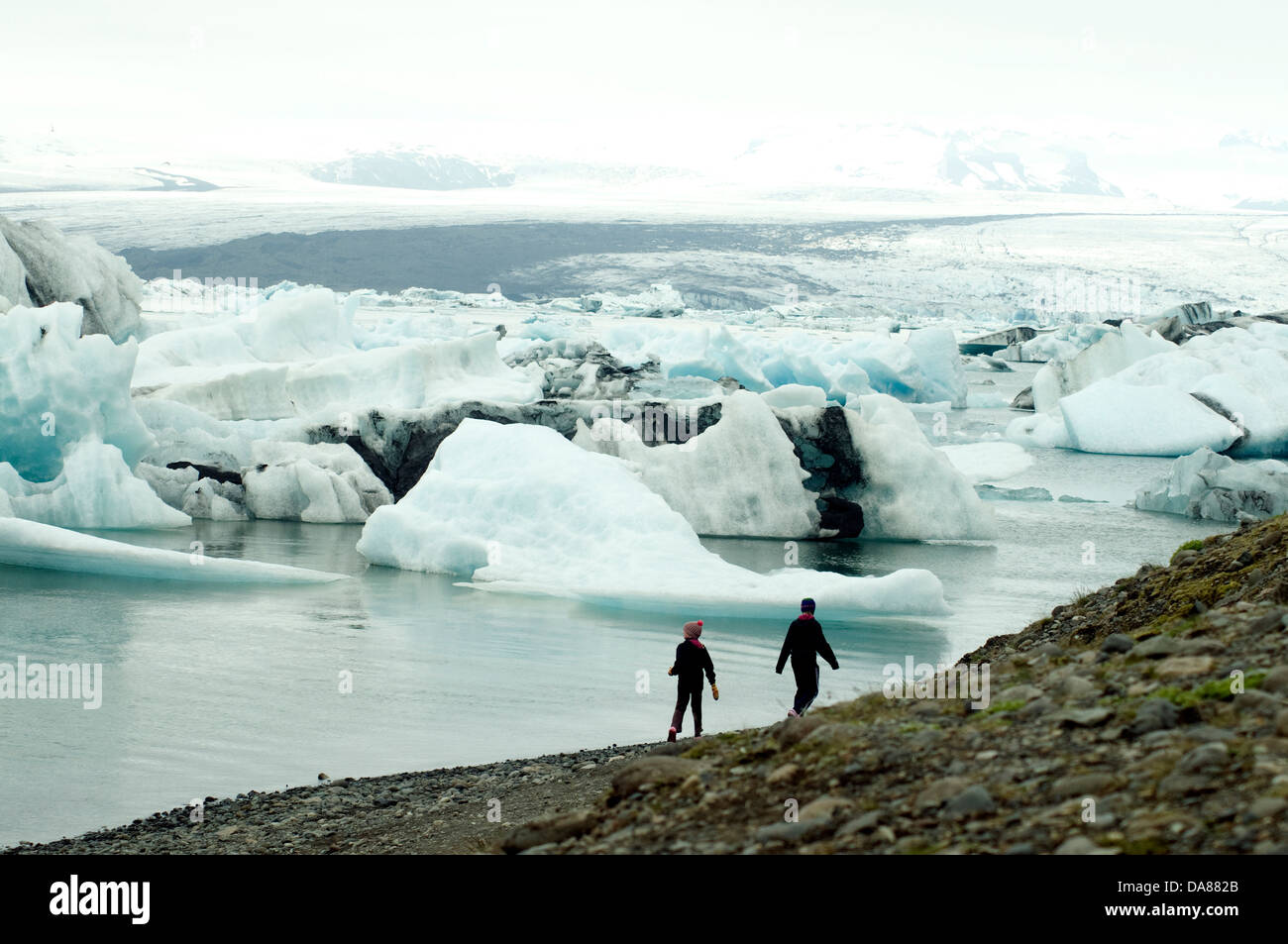 A couple walks the shore or Iceland's Jökulsárlón glacier lagoon taking ...