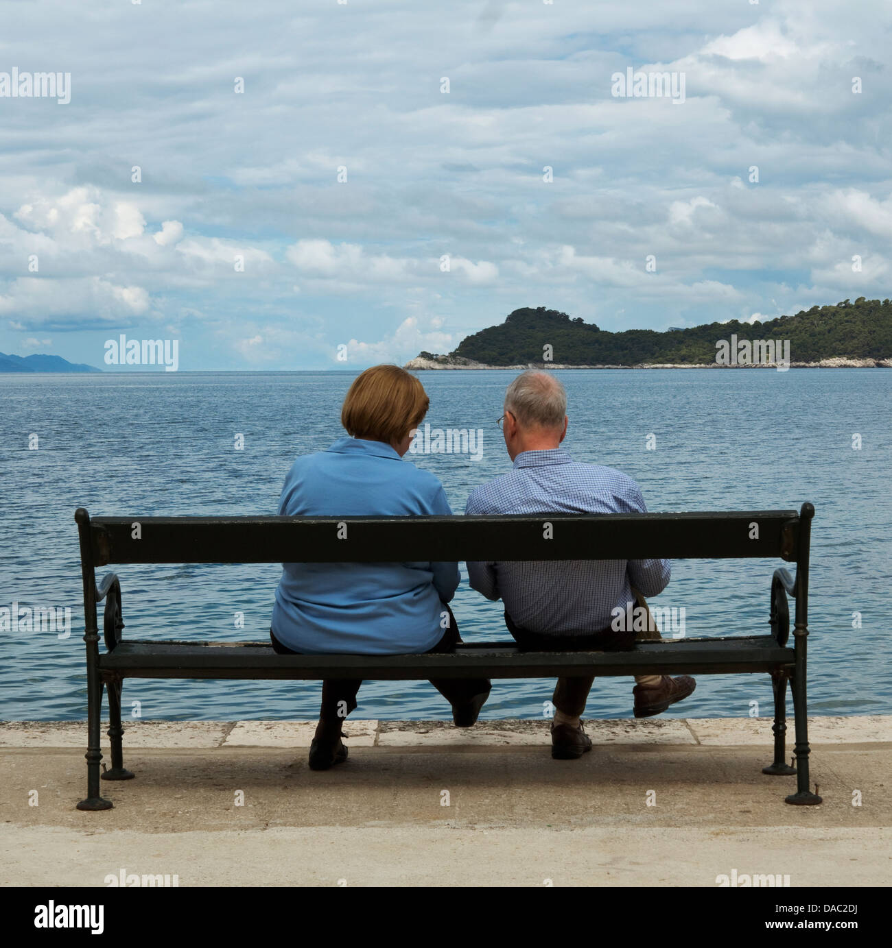 middle aged couple sitting on harbour bench overlooking bay on island ...