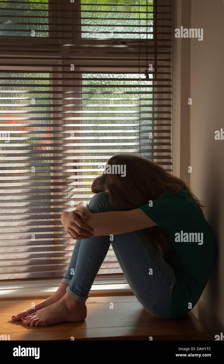 A teenage girl, head between her knees, sitting by a window with light ...