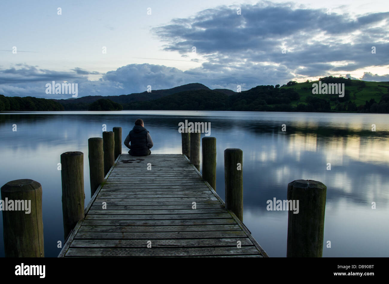 A lonely person is sitting on a wooden pier on a calm lake after sunset ...