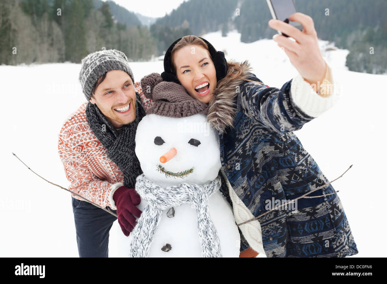 Enthusiastic couple taking self-portrait with snowman in snowy field ...