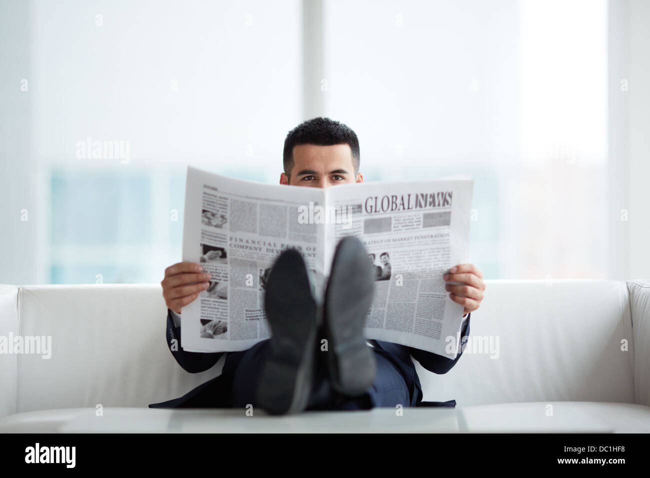 A young businessman on sofa looking at camera while reading newspaper ...