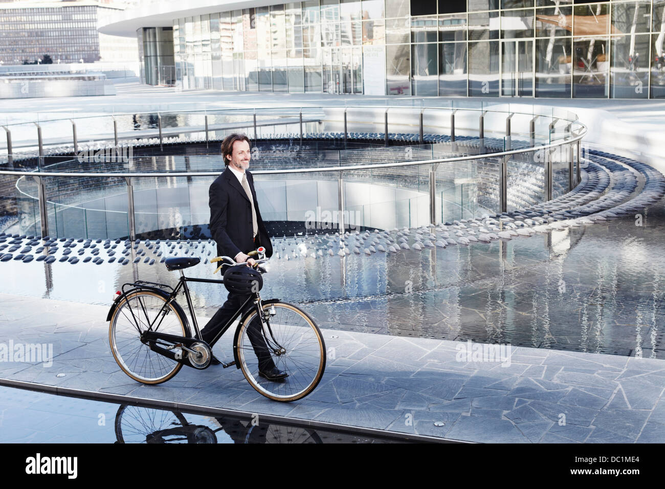 Mid adult businessman walking with bicycle by water feature in city ...