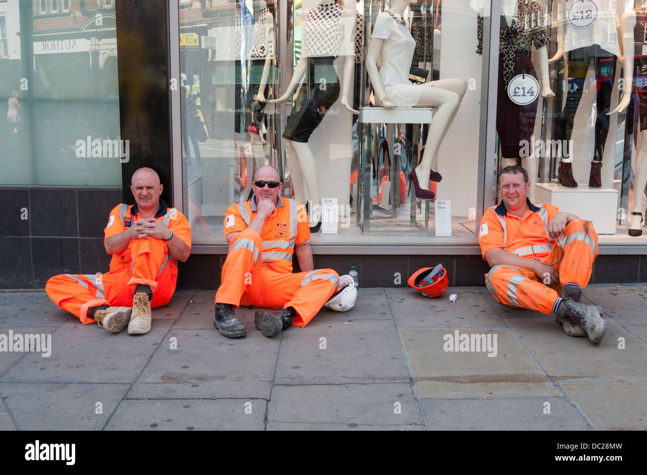 Three workmen in orange Hi Vis clothing take a break outside a shop in ...
