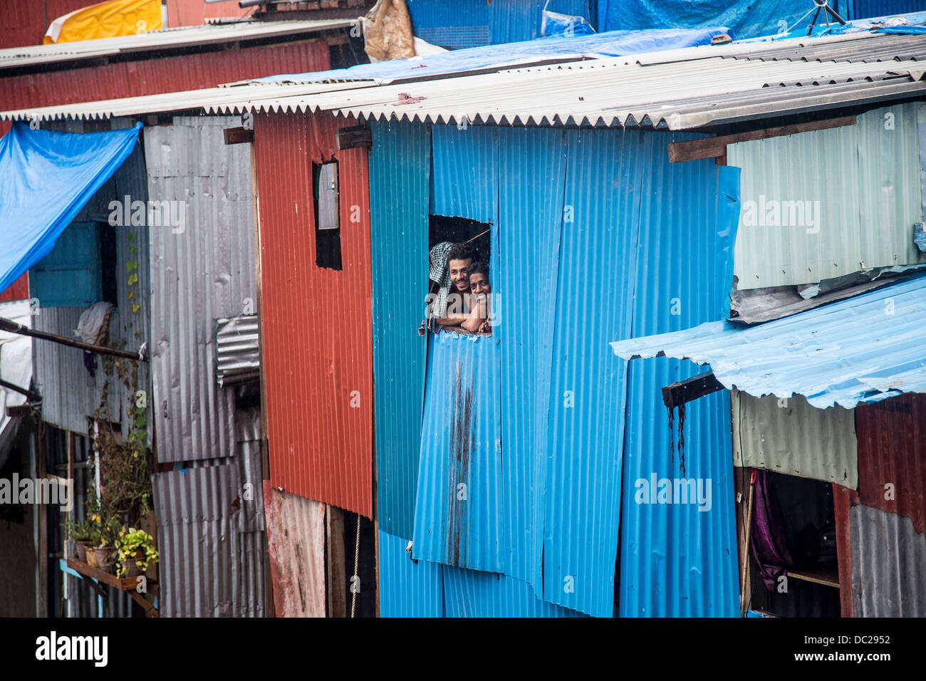 Two smiling men look out of slum window in corrugated iron wall of ...