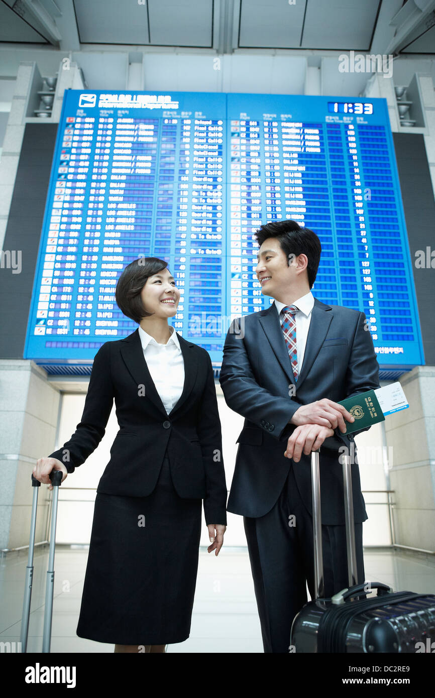 a businessman and woman in front of departure timetable with suitcases ...