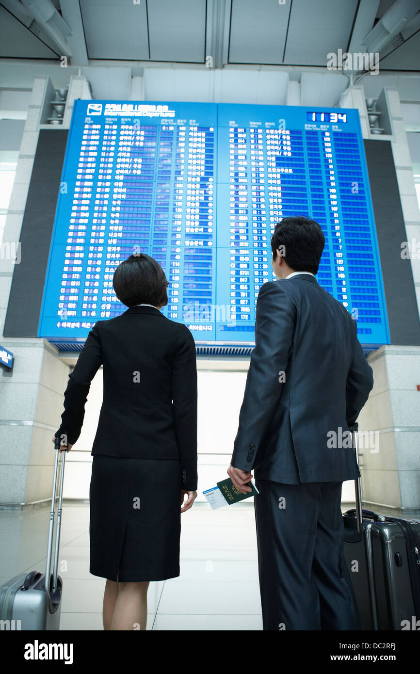 a businessman and woman in front of departure timetable with suitcases ...