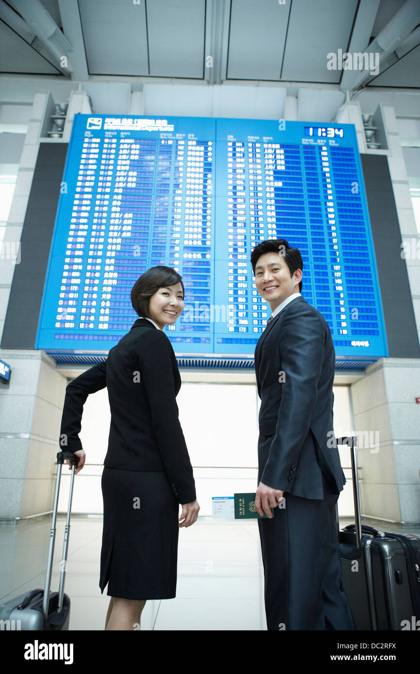 a businessman and woman in front of departure timetable with suitcases ...