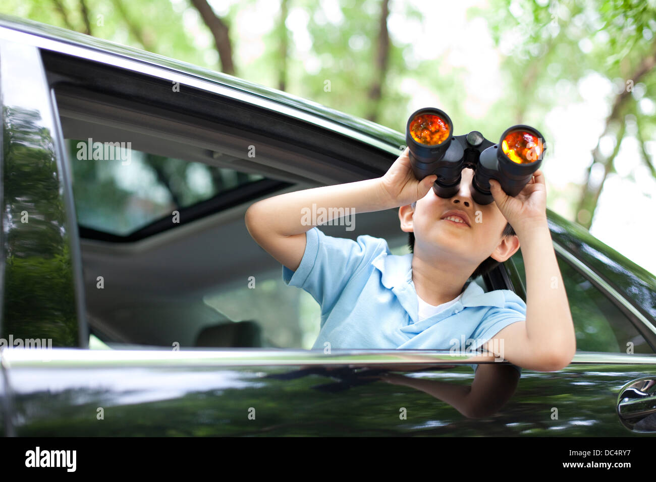 Cheerful boy leaning out of car window and looking with binoculars ...