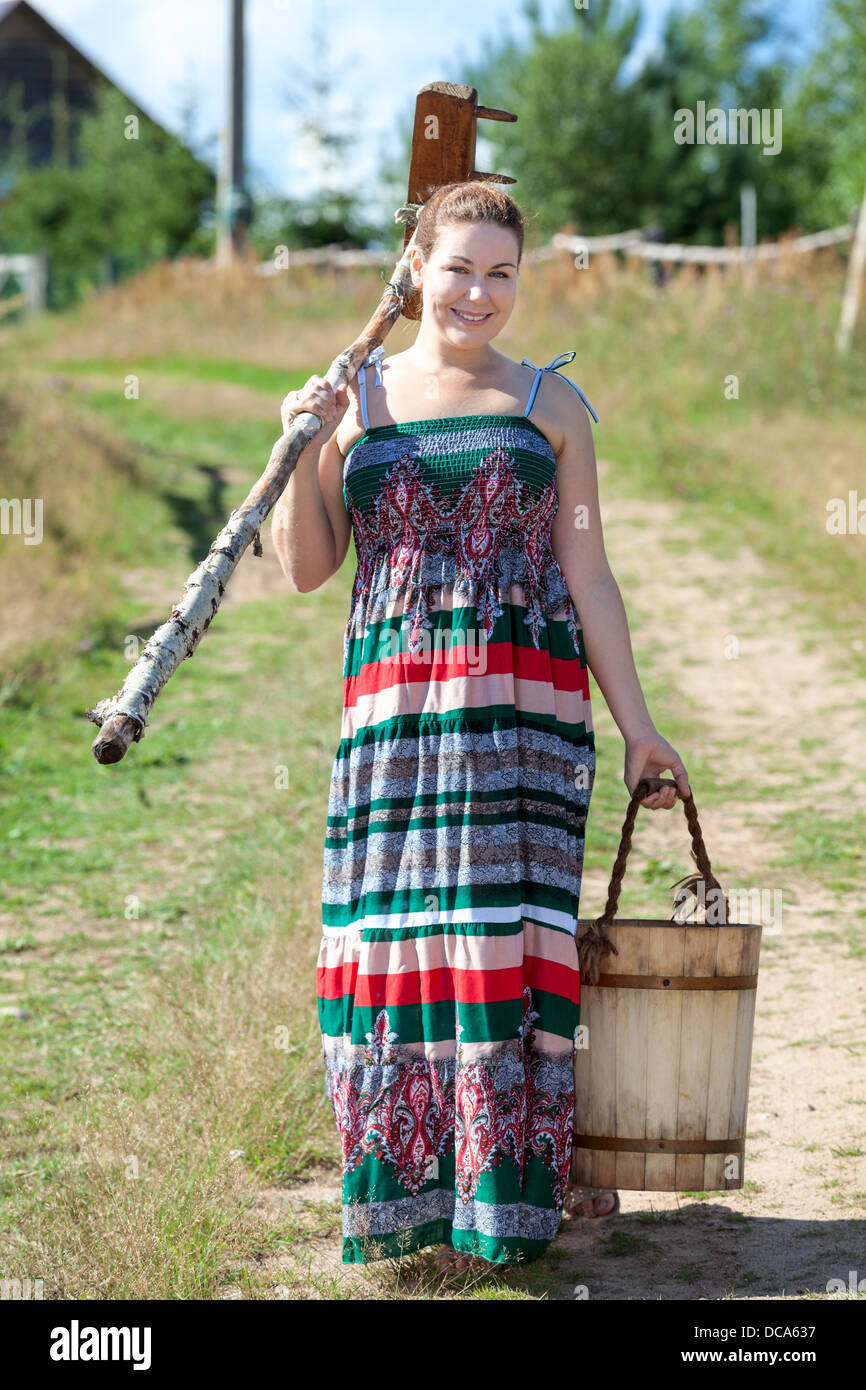 Pretty young village woman with wooden rake and bucket on country road ...