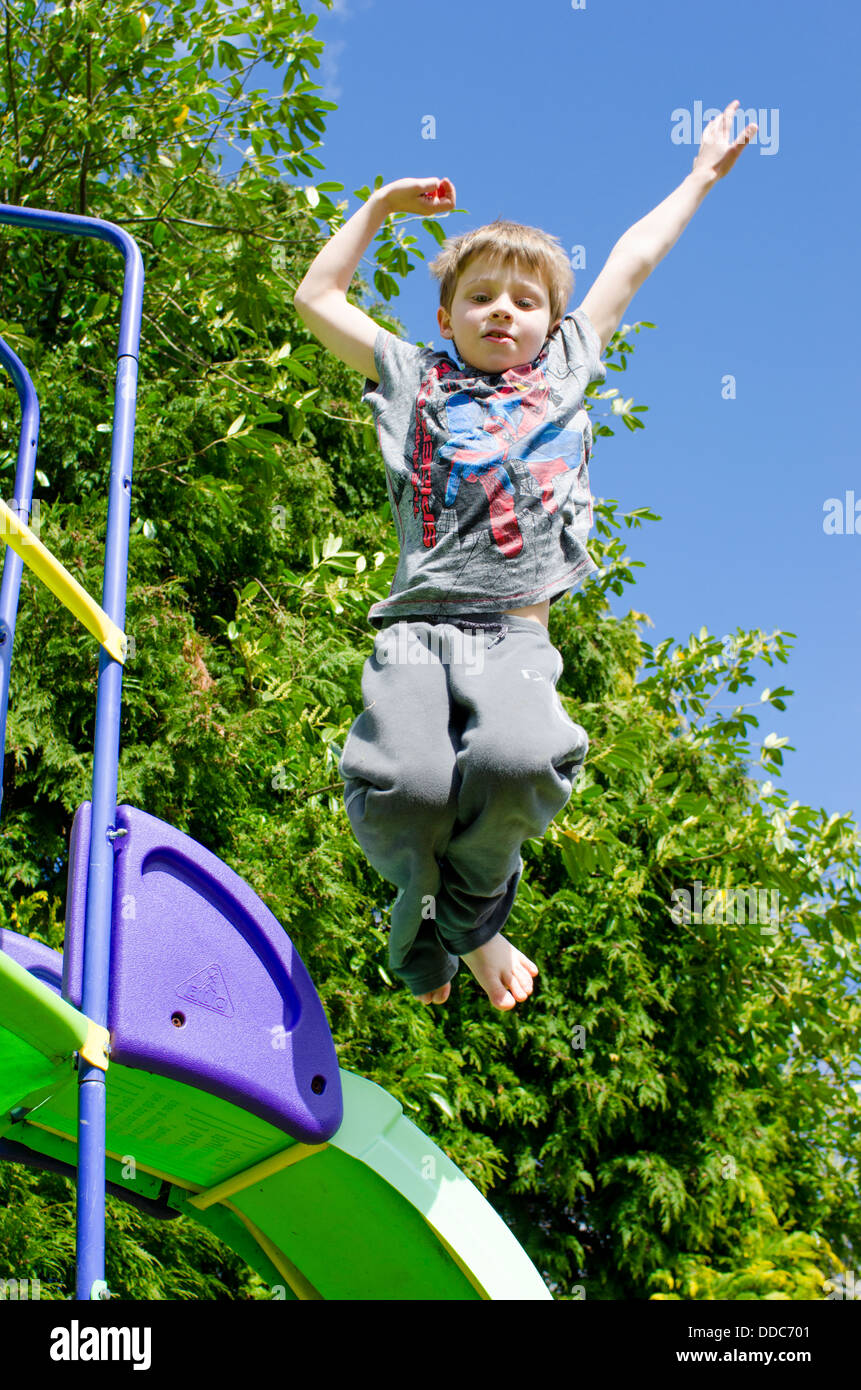 eight year old boy jumping from a climbing frame slide in garden onto a ...