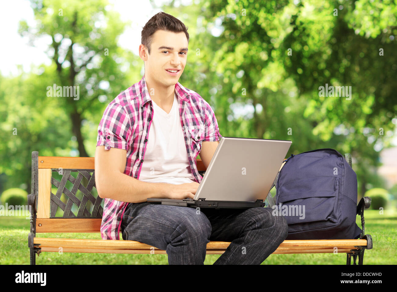 Smiling male student sitting on a wooden bench and working on a laptop ...