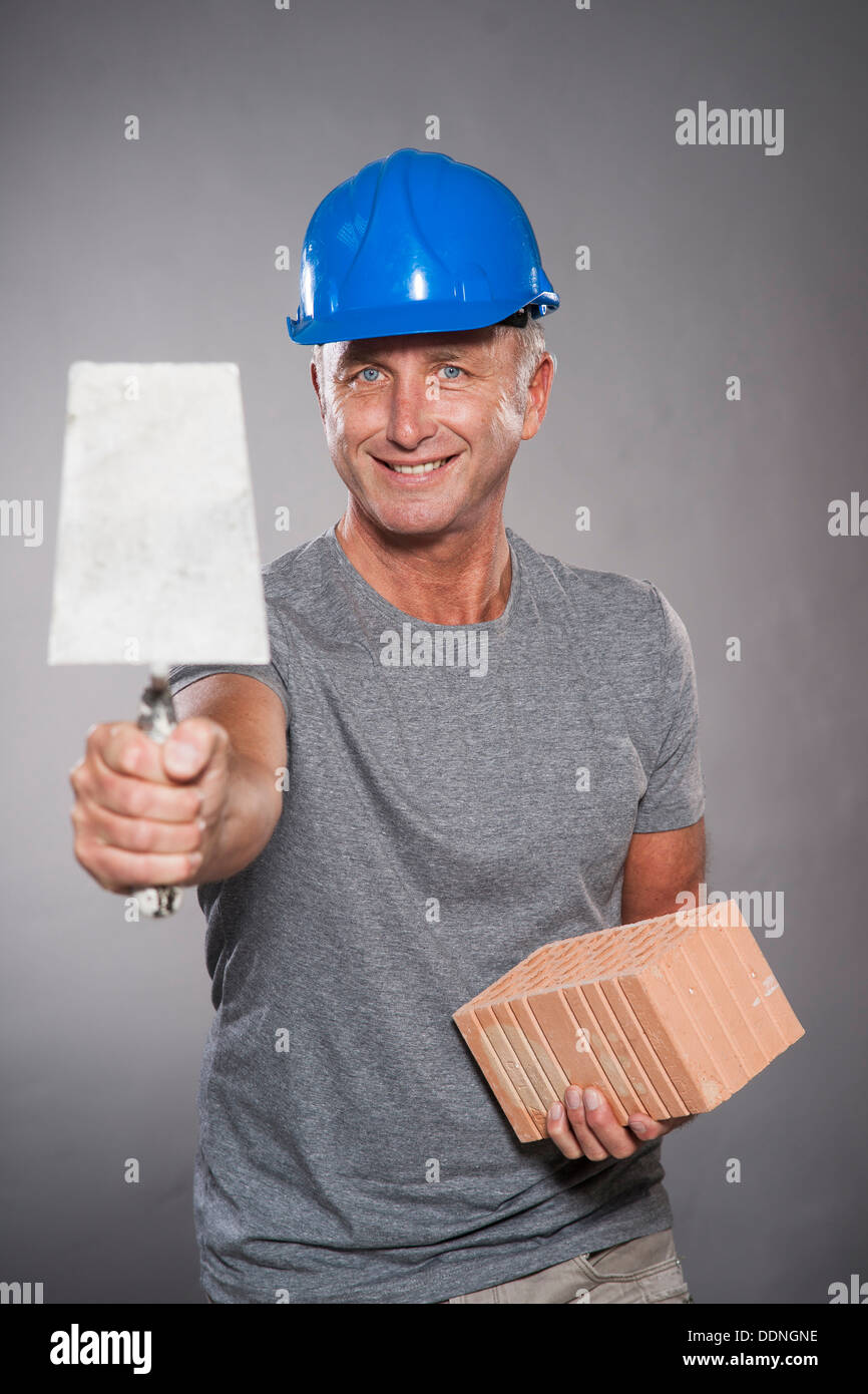 Smiling mature man wearing hard hat, trowel and brick Stock Photo - Alamy