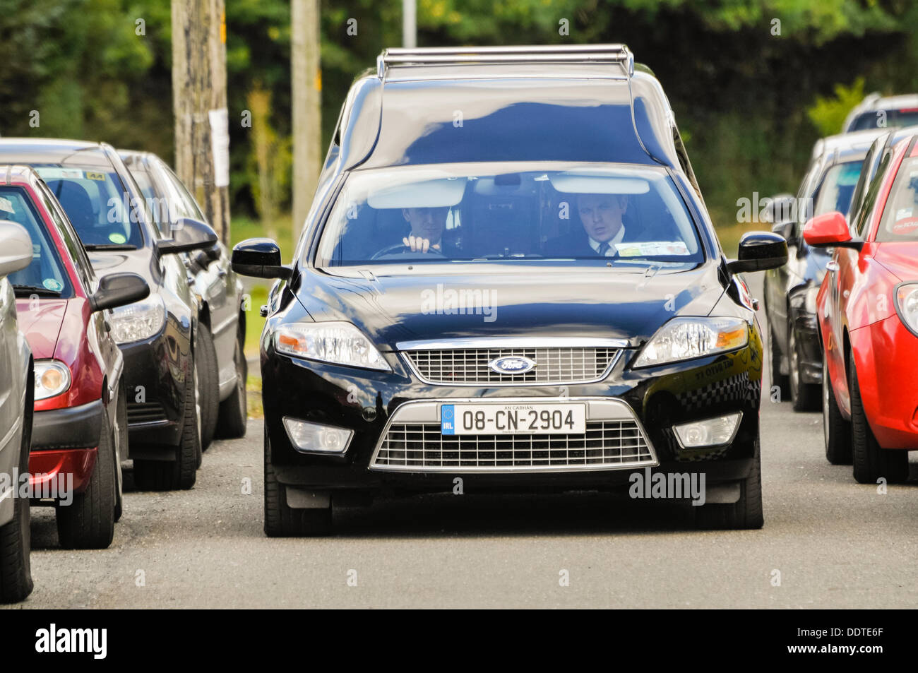 A black Ford hearse drives slowly down a narrow road with cars parked ...