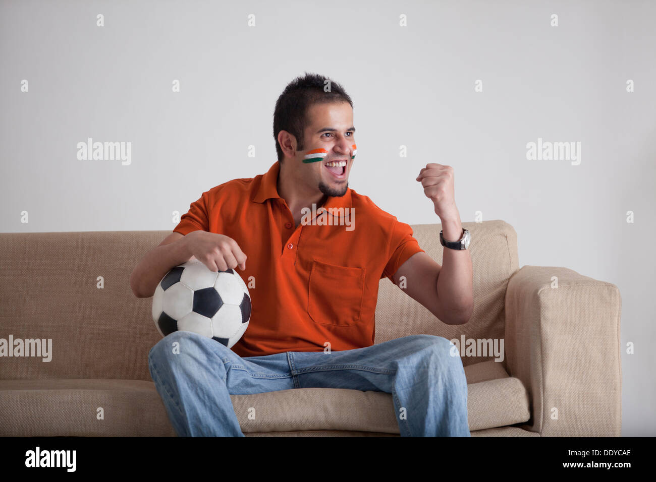 Excited young man with soccer ball cheering while sitting on sofa at ...