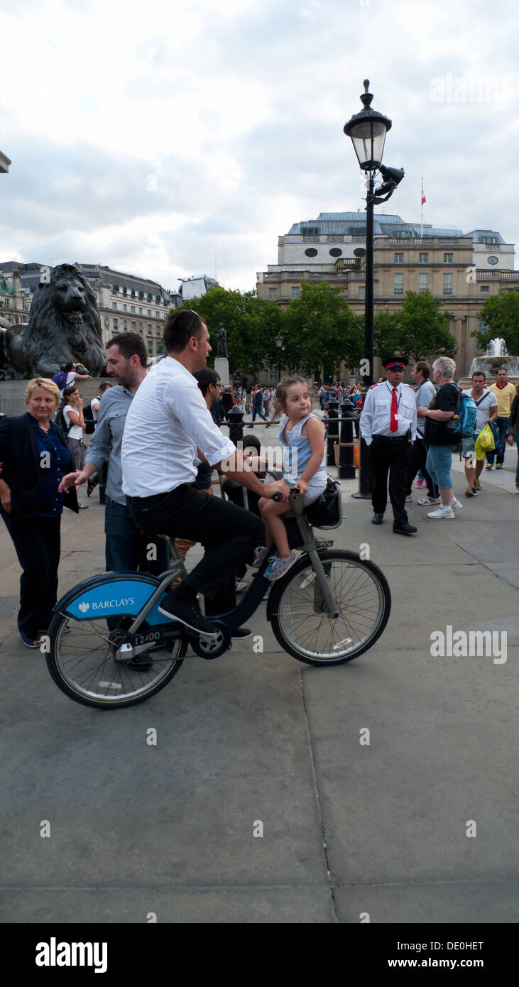 A man riding a Boris bike in Trafalgar Square with a child sitting on the handlebars London ...