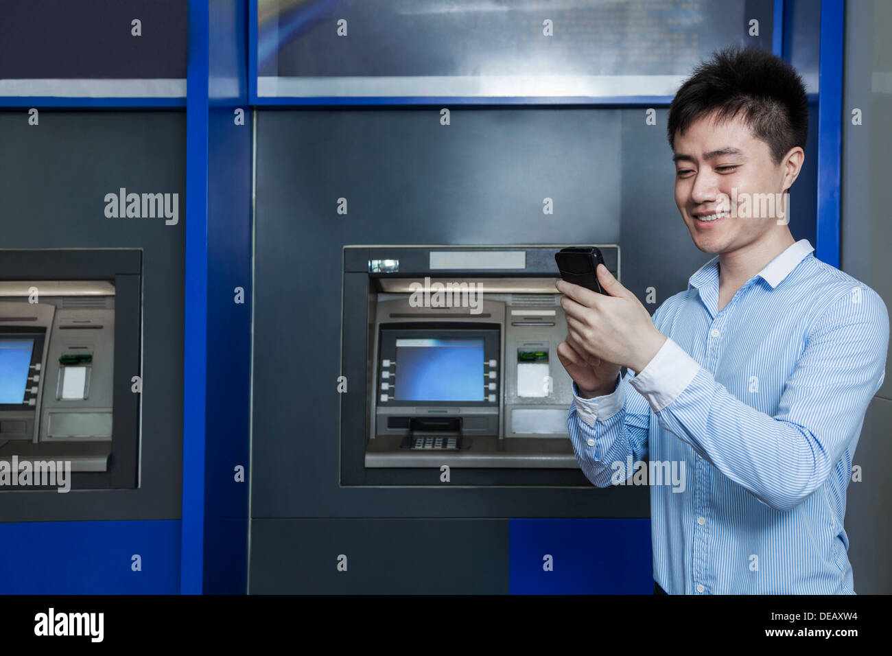 Smiling young businessman standing in front of an ATM and looking at ...