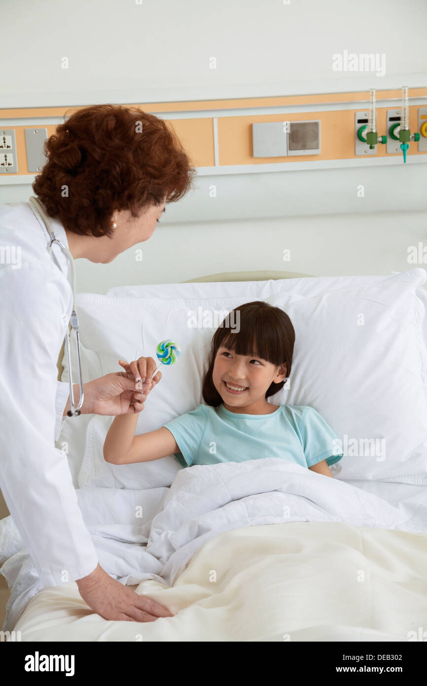 Female doctor giving a lollipop to a young girl lying in a hospital bed ...
