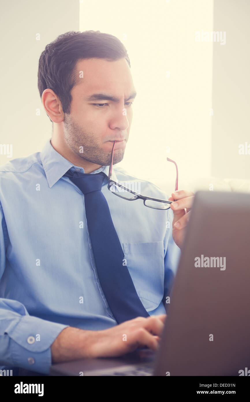 Frowning man biting his eyeglasses and using a laptop Stock Photo - Alamy
