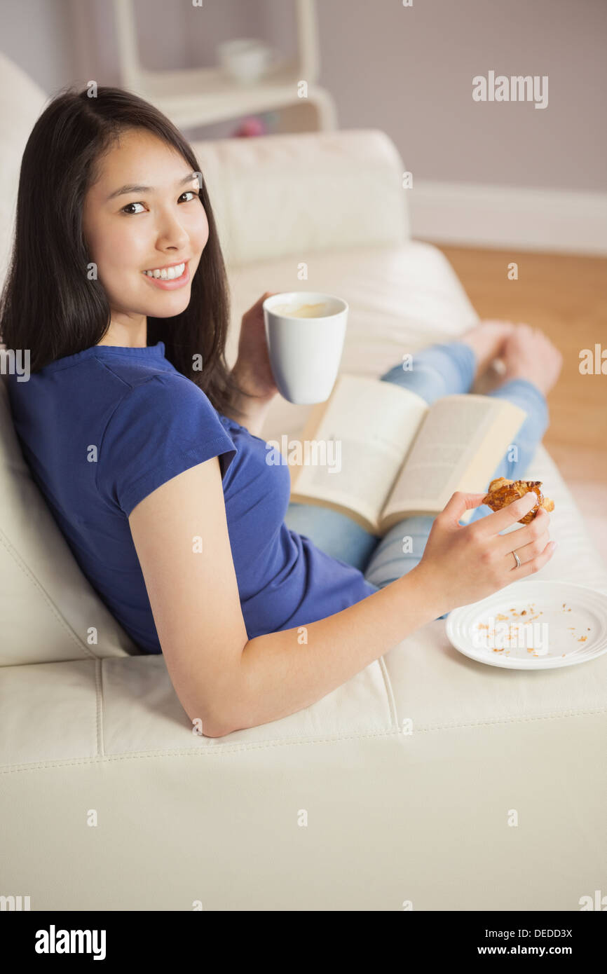 Young woman reading a book and eating pastry with coffee turning to the ...