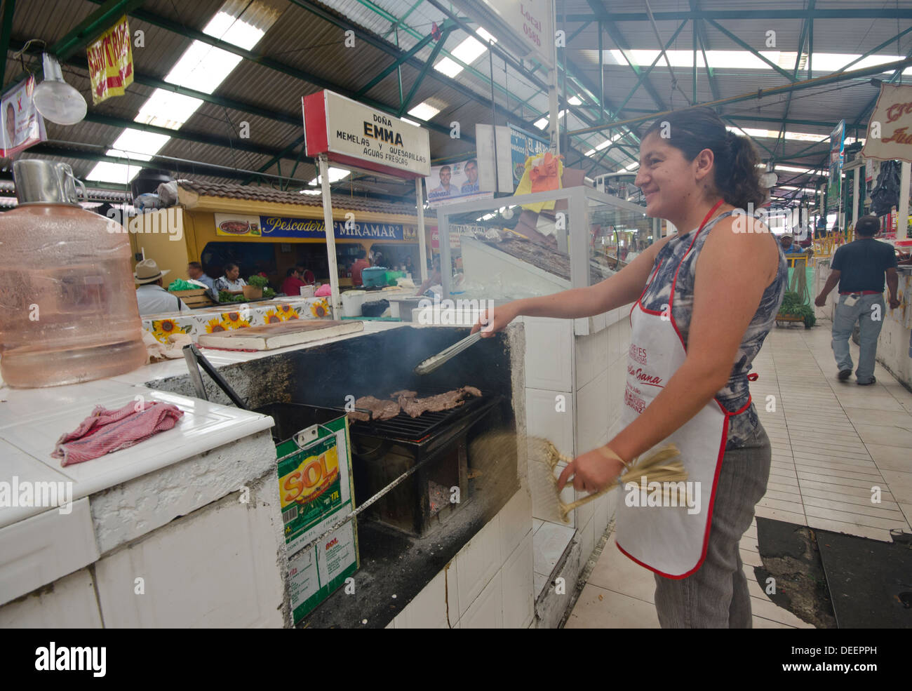 Woman fanning the fire of a brazier whilst grilling steaks in a market ...