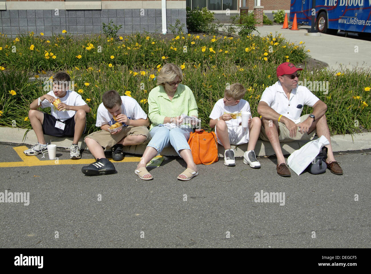 Family relaxes on the curb of a street eating lunch while attending a ...