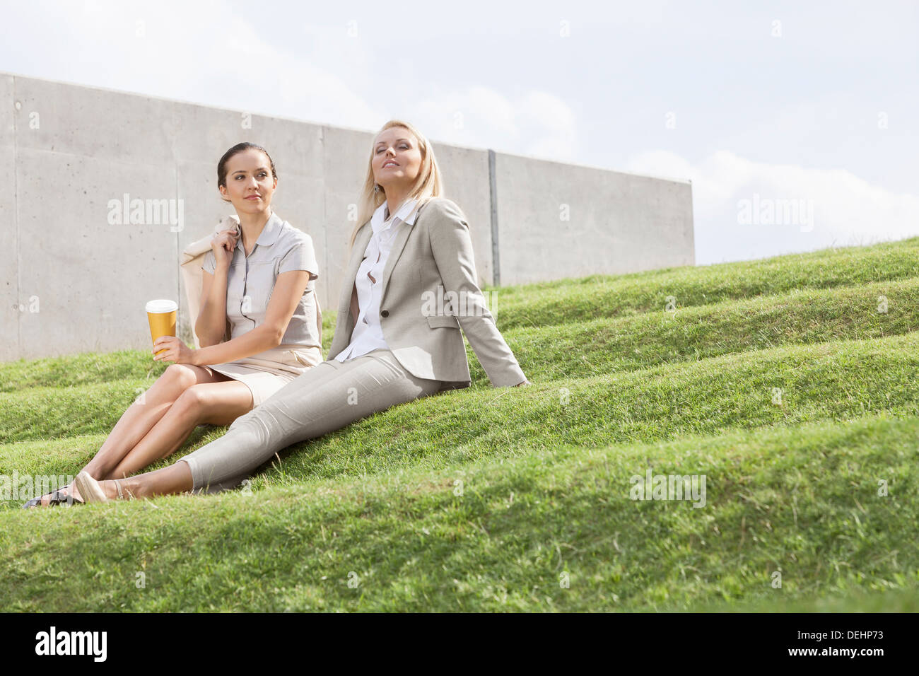 Full length relaxed businesswomen sitting on grass steps against sky ...