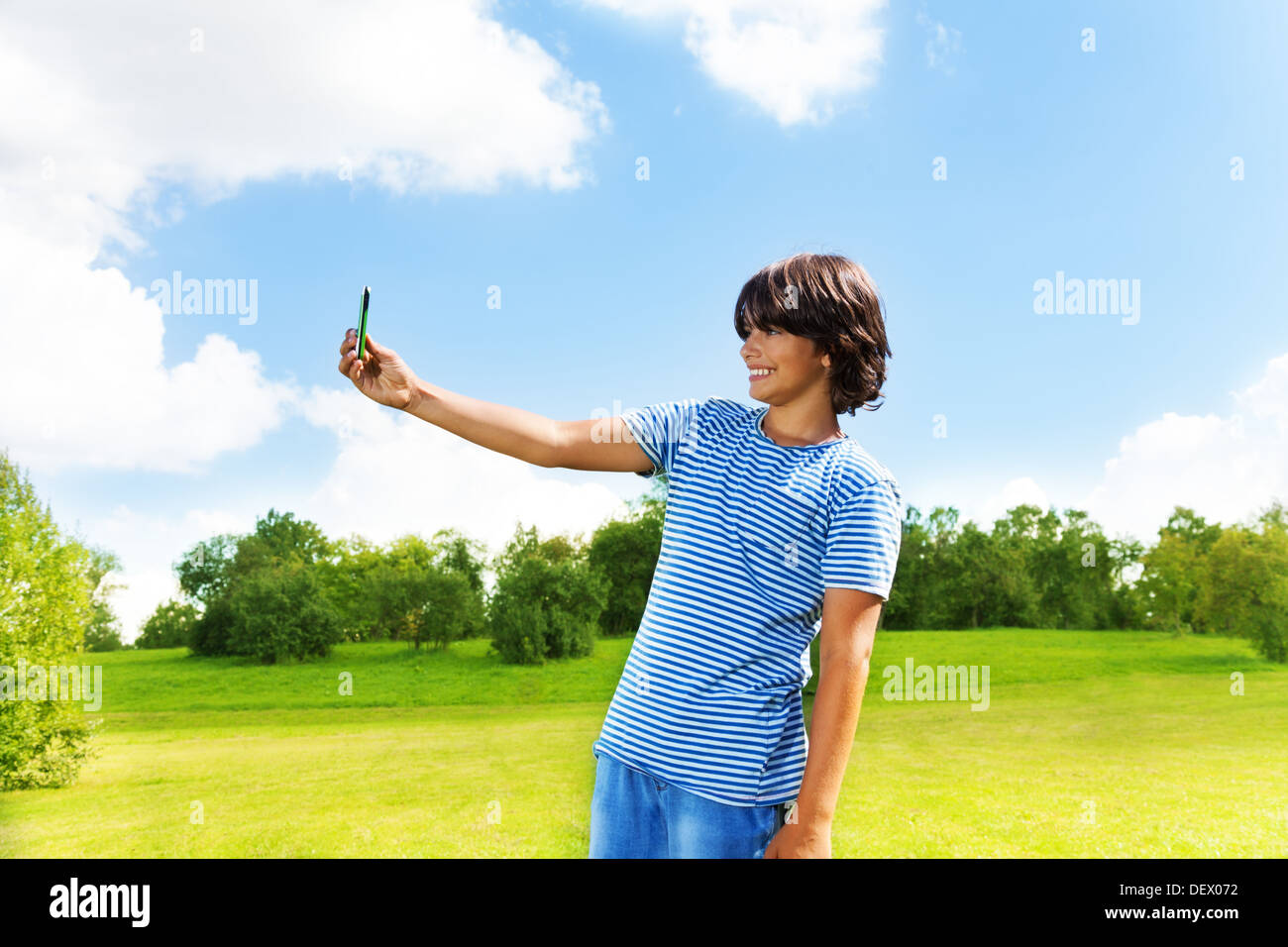 Happy smiling teen boy taking a picture of himself with camera on the cell phone in the park on ...