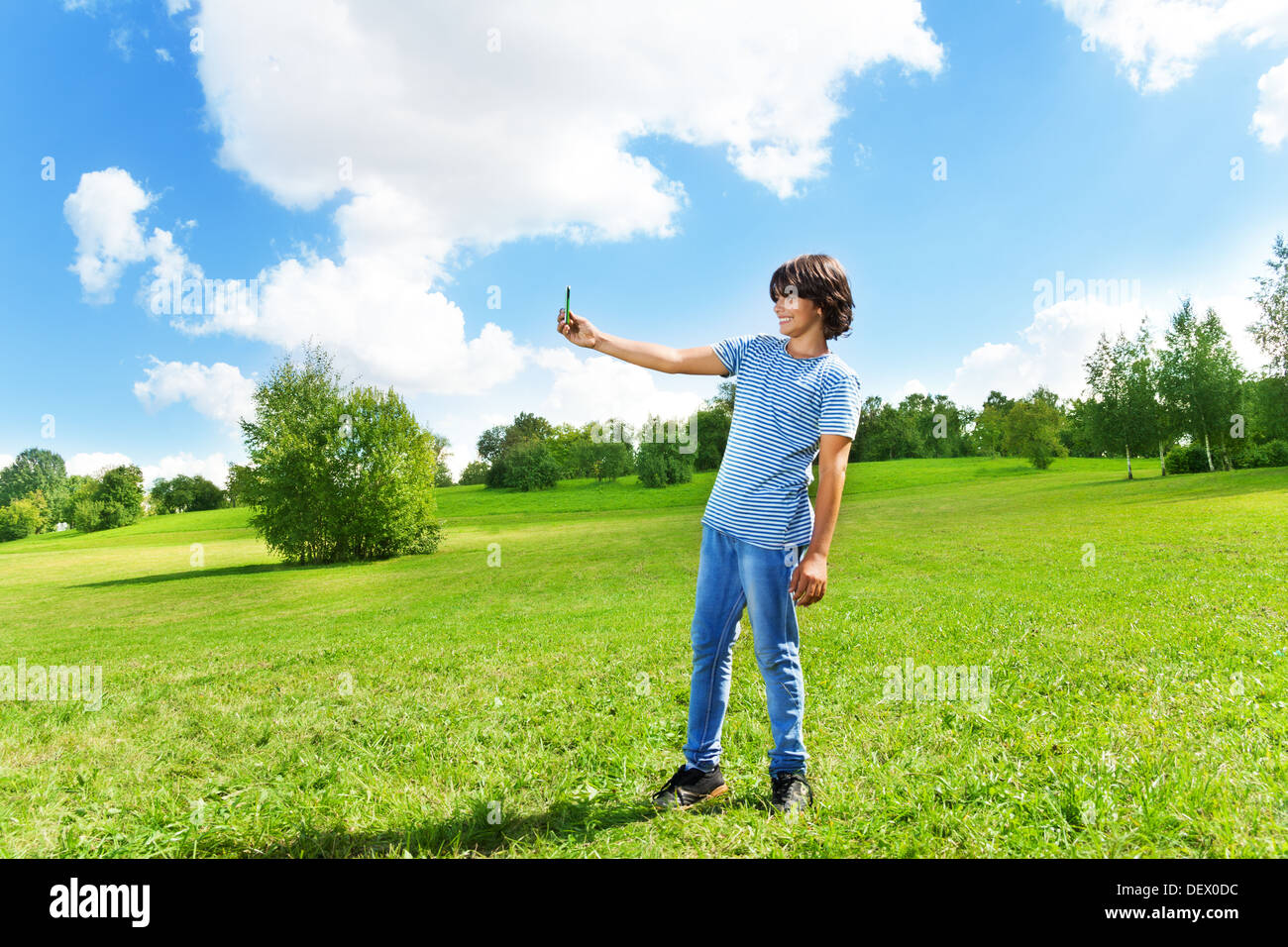 Happy smiling teen boy taking a picture of himself with smartphone in the park on sunny summer ...