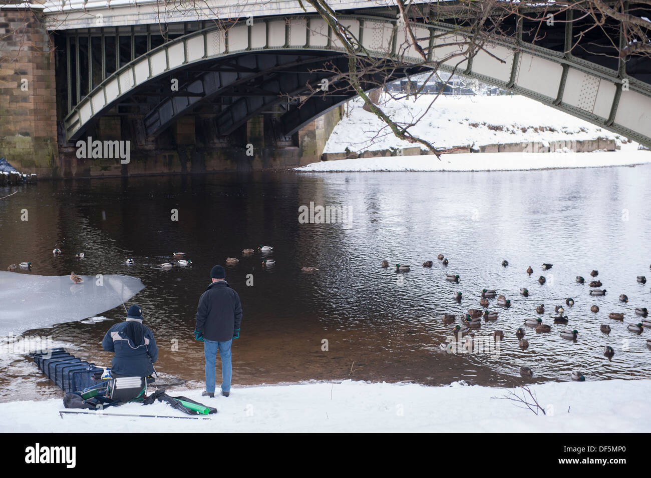 2 men in snow by riverbank of partly frozen River Wharfe (1 man sitting ...