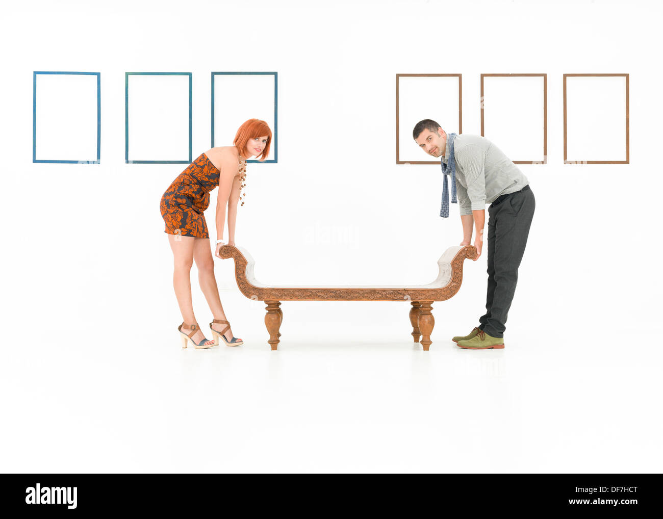 two caucasian people trying to lift up a wooden bench in a white room ...
