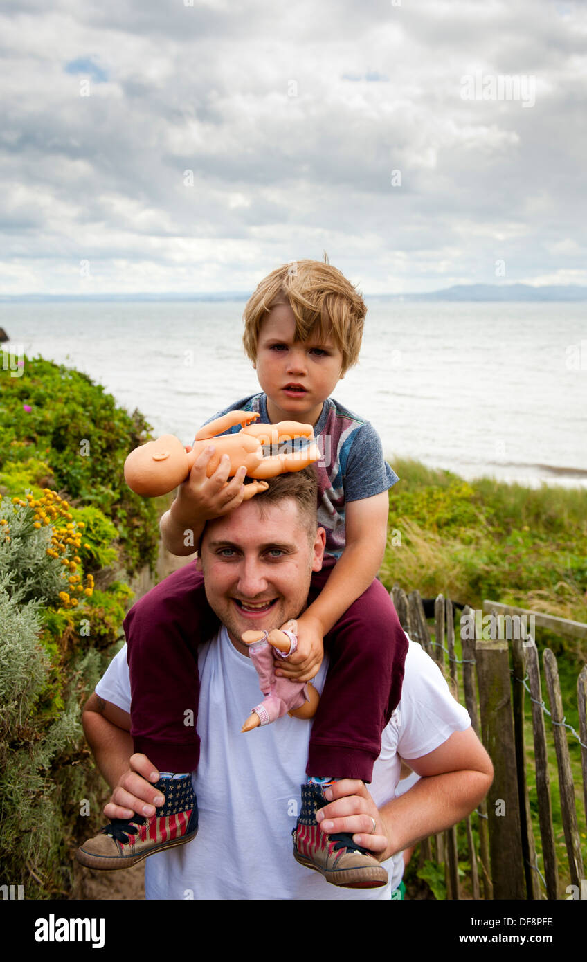 Dad with young son on shoulders, holding a doll with beach view at ...