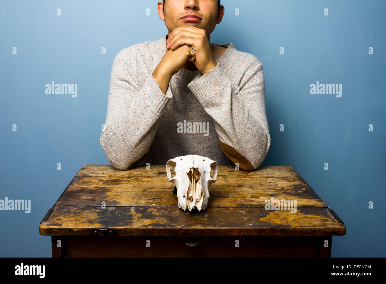 YOung man is sitting at a desk with a goat's skull in front of him ...