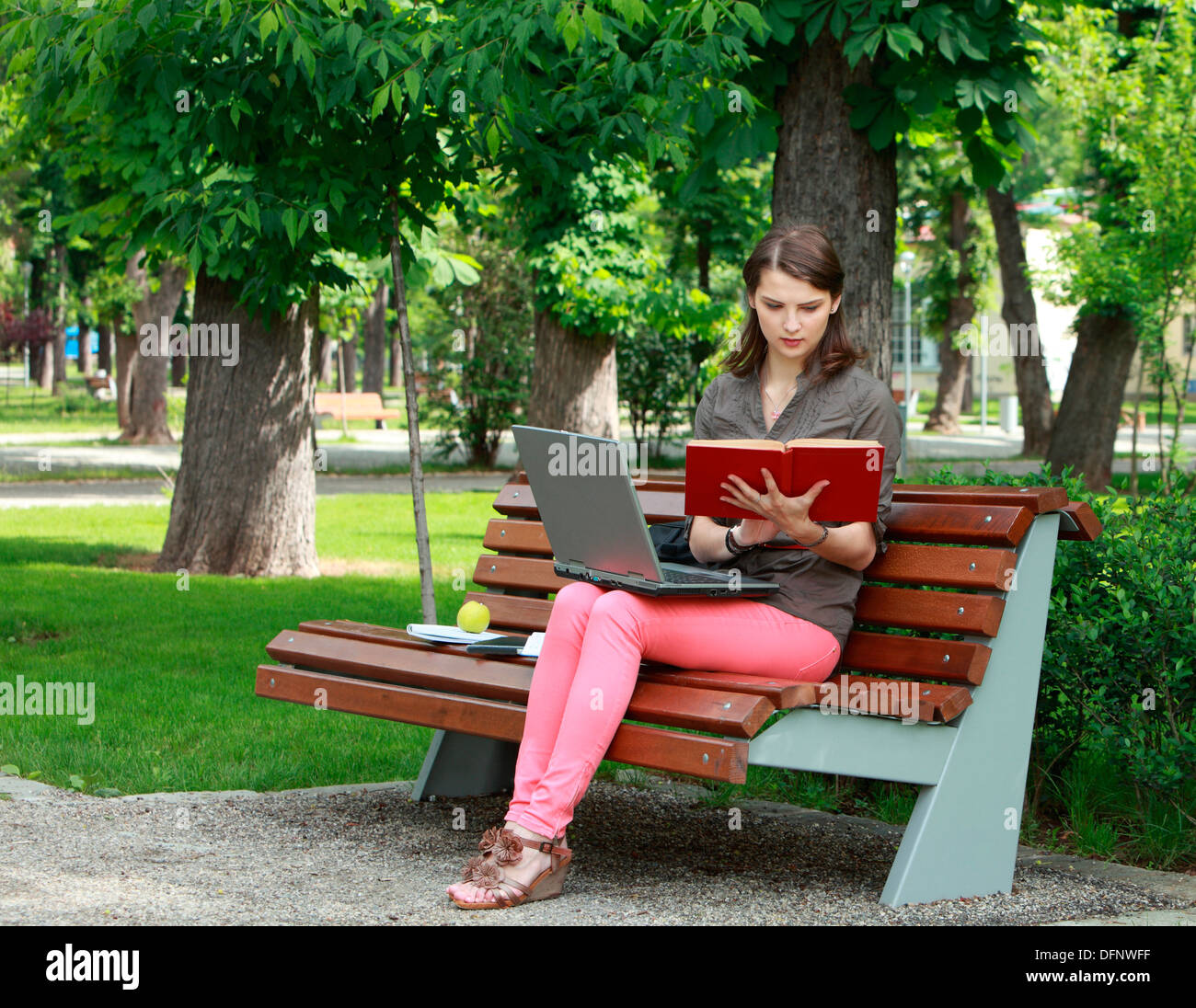 Young woman with a laptop reading a book and sitting on a bench outside ...