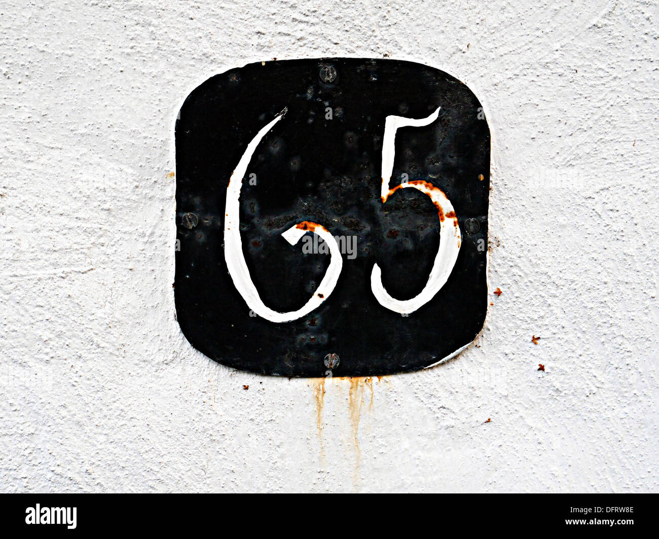 Old black rusty sign showing the number of 65 against a white wall on a ...