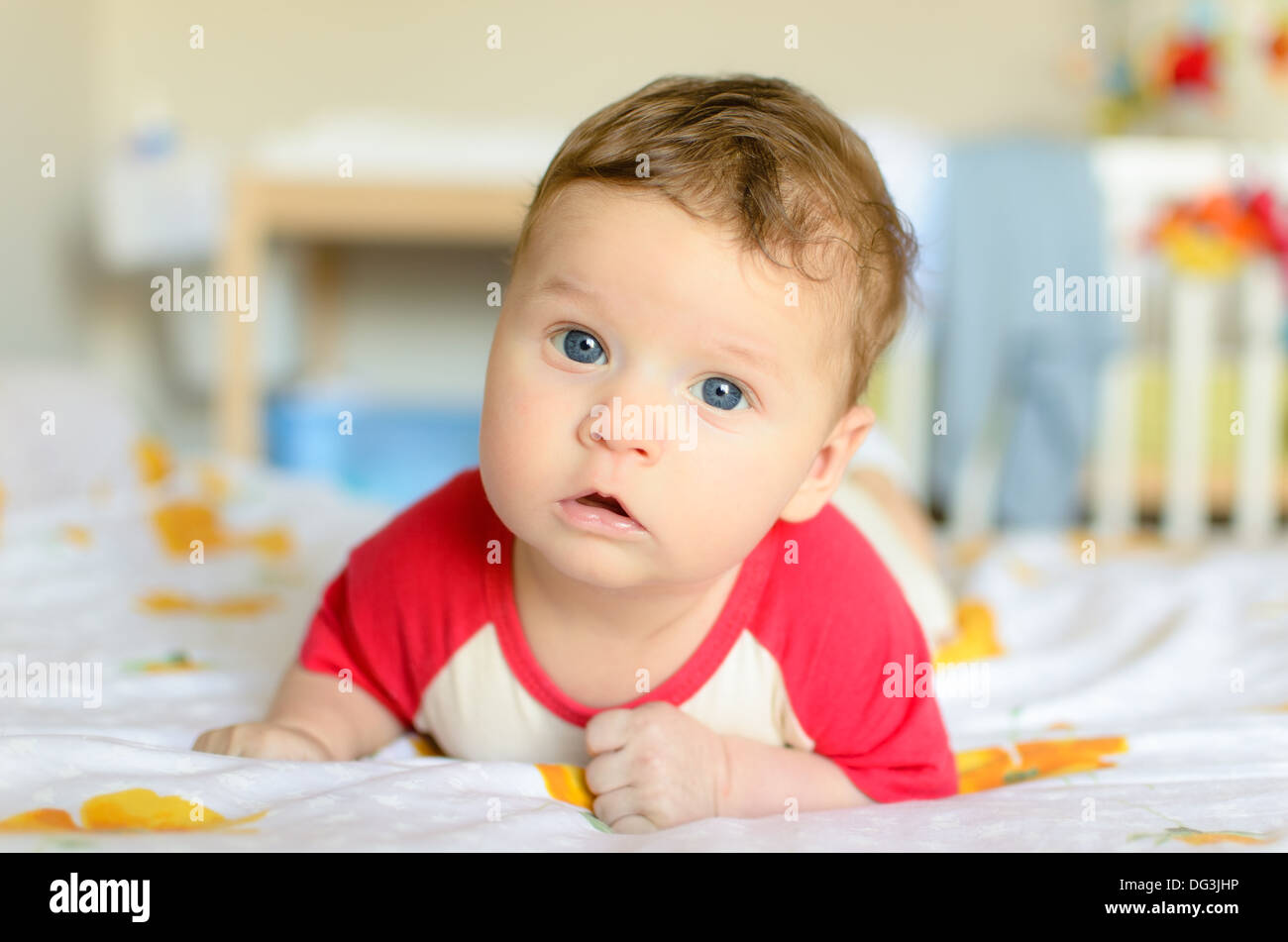 Cute serious baby lies on his tummy in nursery and looks at camera ...