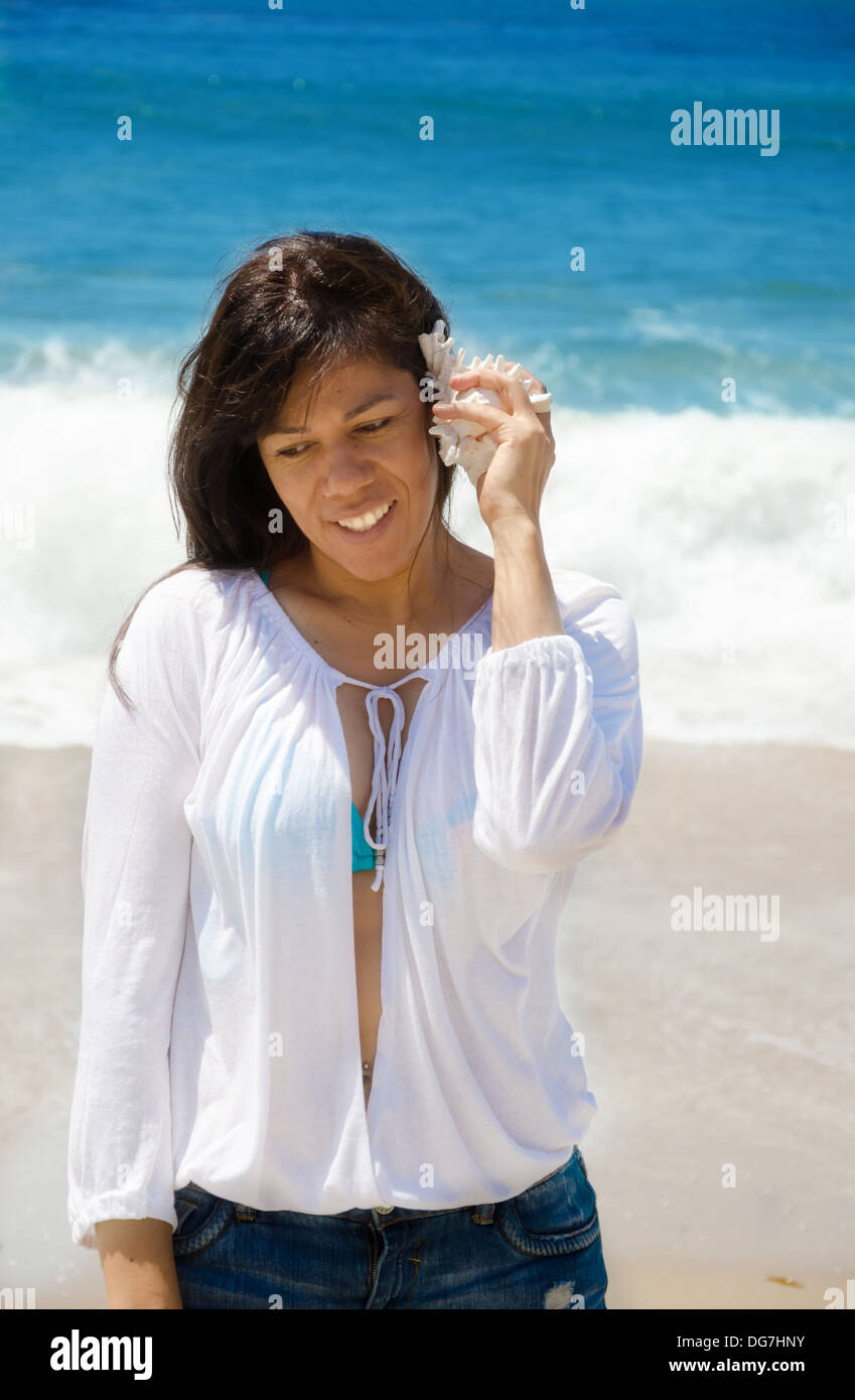 Beautiful hispanic woman hearing the sounds of seashell by the ocean ...