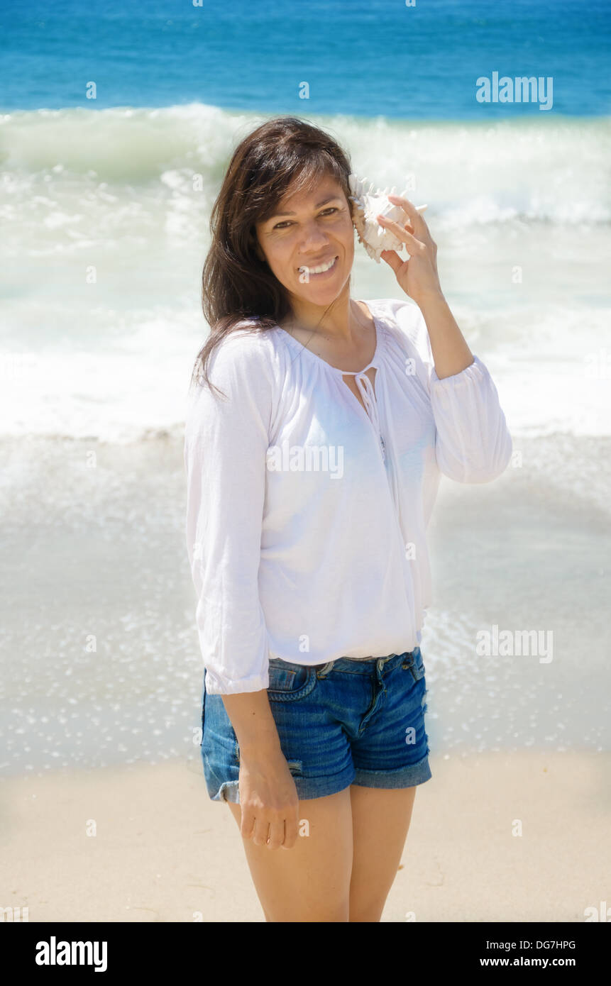 Beautiful hispanic woman hearing the sounds of seashell by the ocean ...